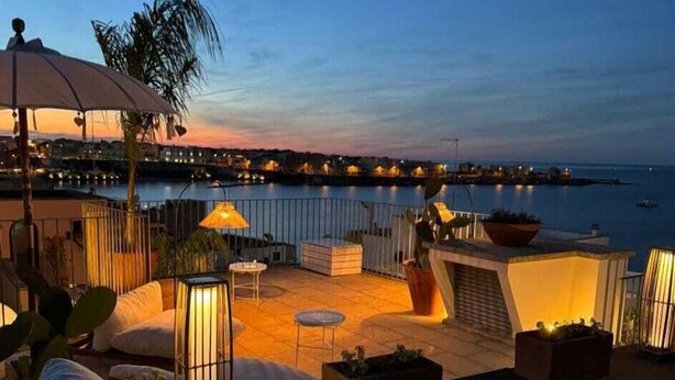 Terraço no telhado com vista para a orla ao entardecer, com área de estar ao ar livre e iluminação ambiente.