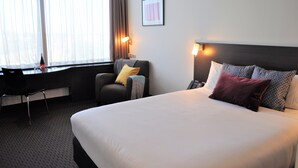 Quarto de hotel com cama, escrivaninha, cadeira e janela com cortinas.