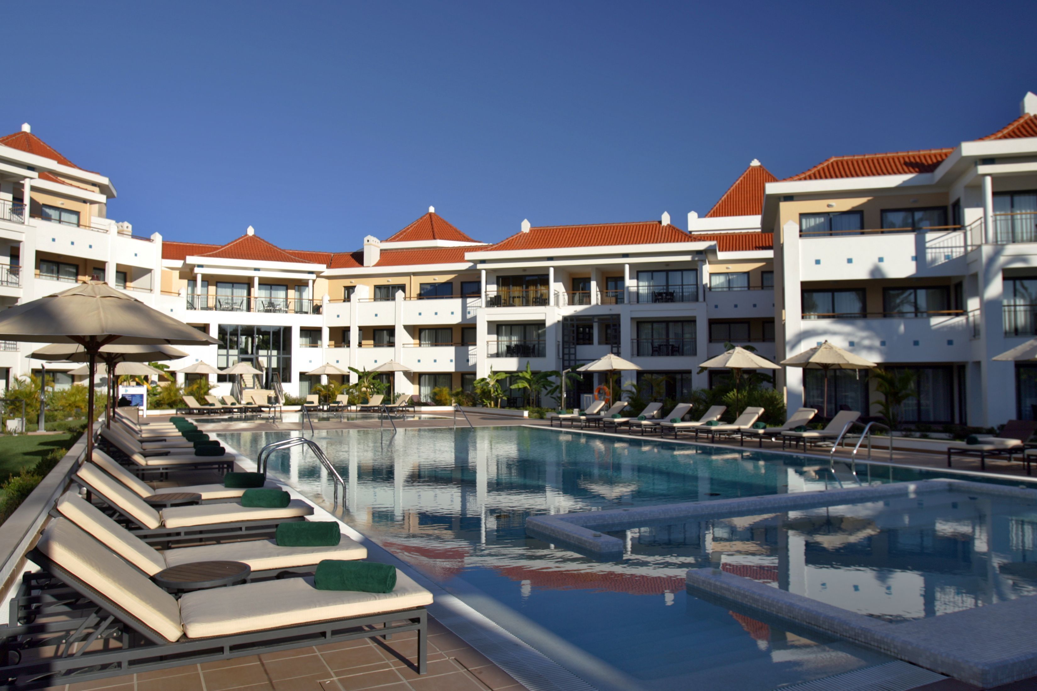 Área de piscina de hotel com espreguiçadeiras e guarda-sóis, rodeada por edifícios altos com varandas.