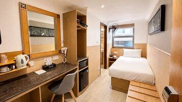 Quarto de hotel compacto com cama, escrivaninha com telefone, espelho e janela.