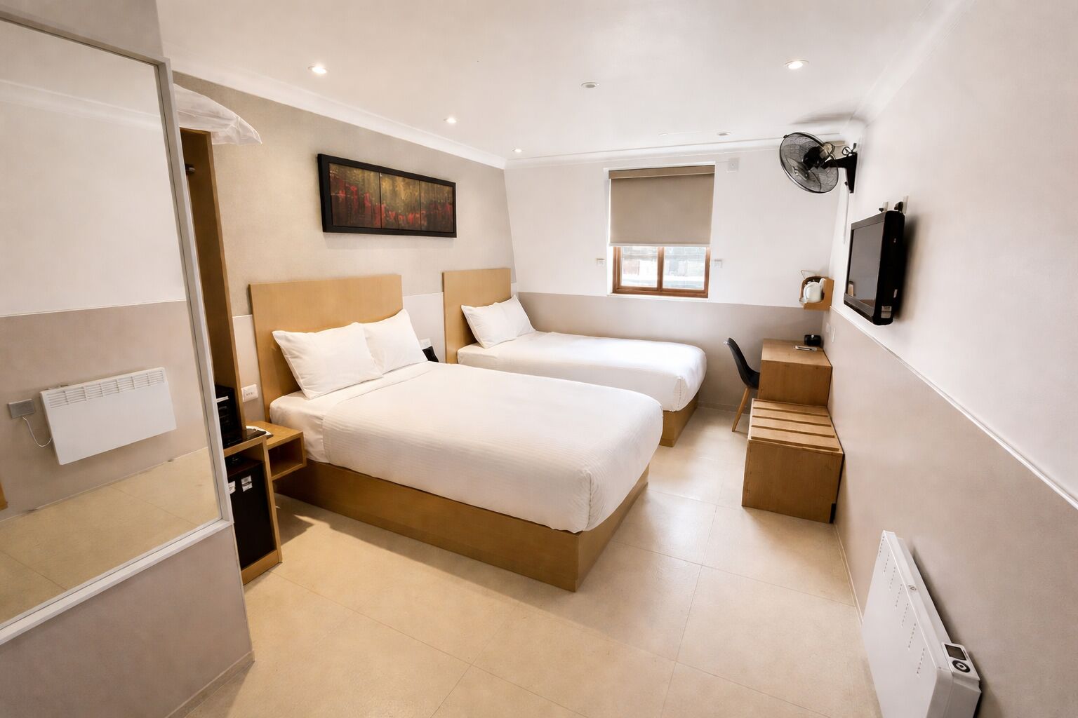 Quarto de hotel com duas camas, uma escrivaninha, uma cadeira, um radiador, uma televisão e uma janela com vista.