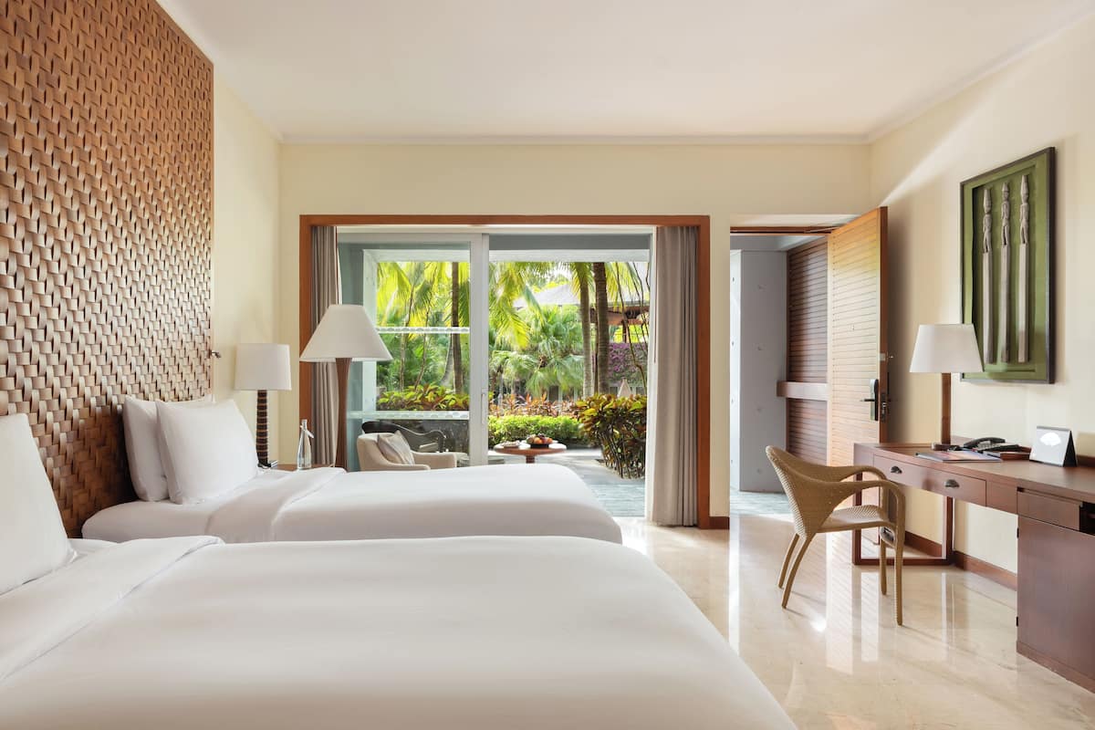 Quarto de hotel com duas camas, uma escrivaninha e vista para um jardim tropical.