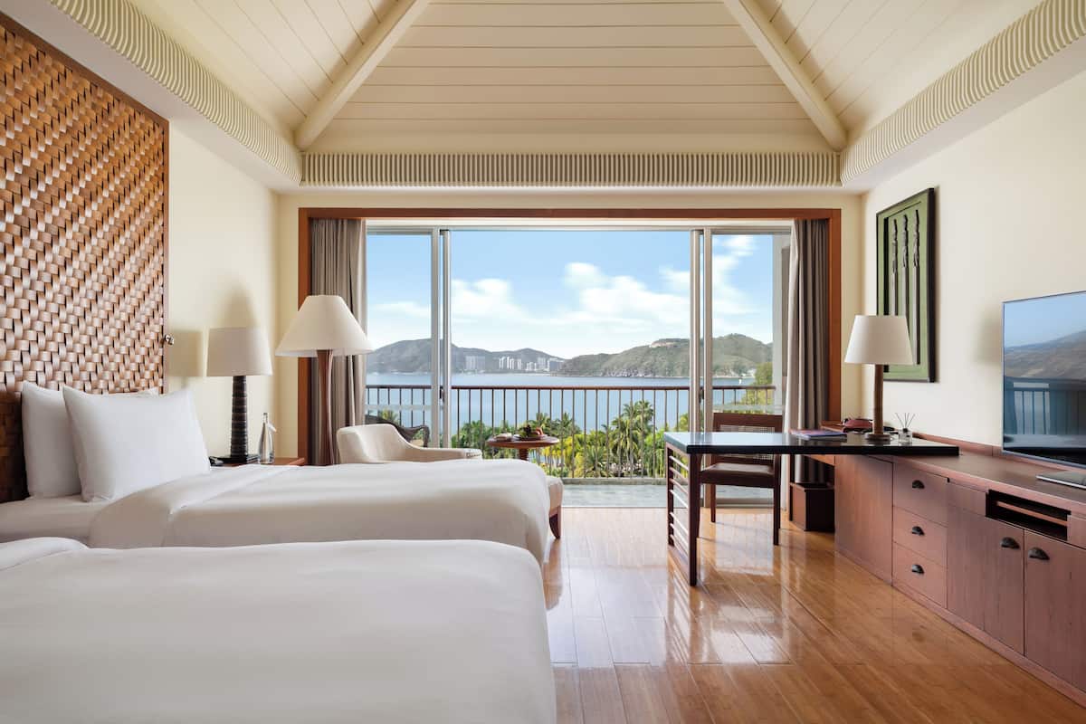 Quarto de hotel com duas camas, uma escrivaninha, televisão e varanda com vista para o mar e montanhas.