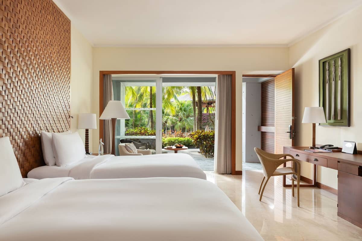 Quarto de hotel com duas camas, uma escrivaninha e vista para um jardim tropical.