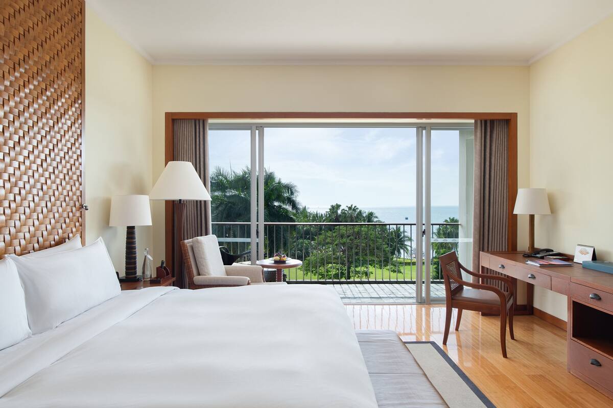 Quarto de hotel com cama grande, escrivaninha, cadeira, mesinha pequena e varanda com vista para o oceano e palmeiras.