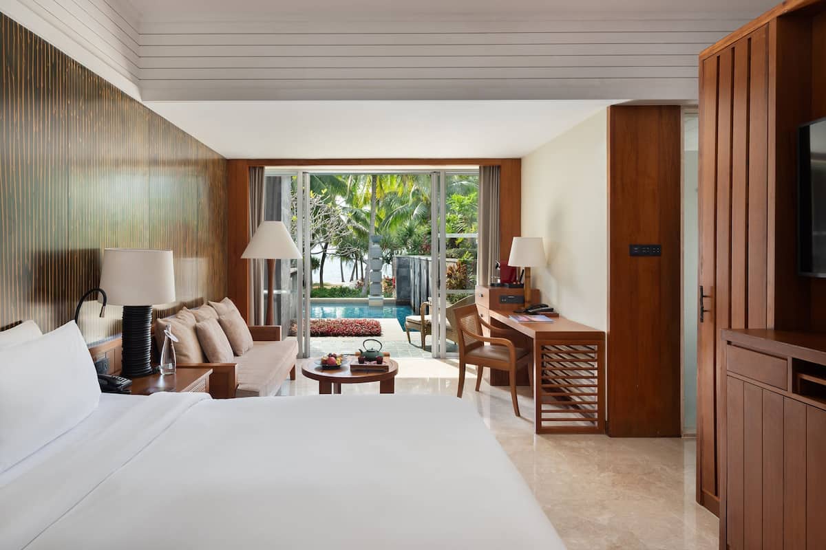 Quarto de hotel com cama, escrivaninha, cadeira, TV e vista para uma piscina e palmeiras.
