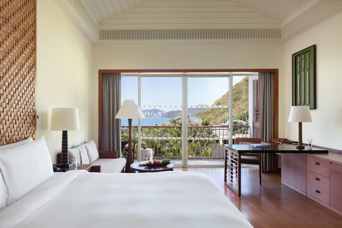 Quarto de hotel com uma cama grande, uma escrivaninha, uma cadeira, uma mesinha com fruta e uma varanda com vista para o mar e colinas.