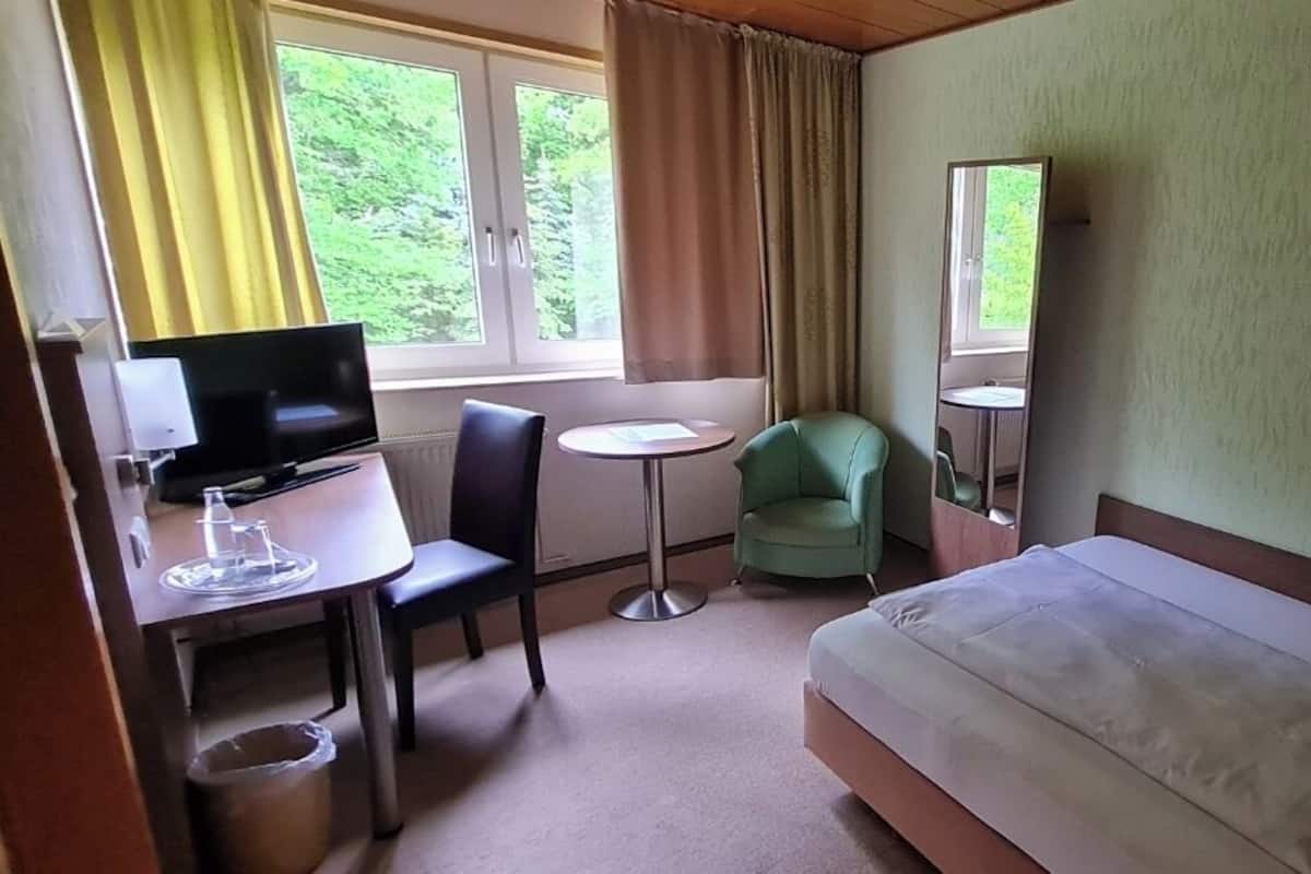 Quarto de hotel com cama, escrivaninha, televisão, cadeira, mesa pequena e janela com cortinas.