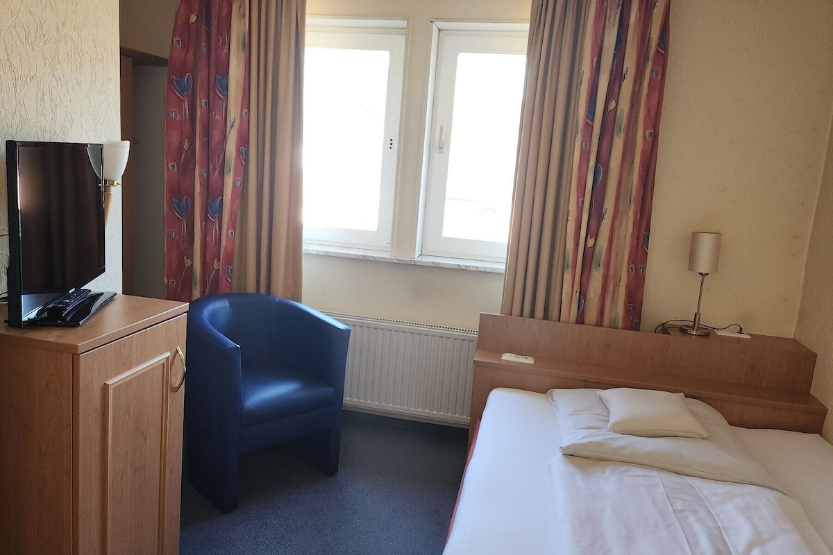 Quarto de hotel com cama, escrivaninha com computador, cadeira e janela com cortinas.
