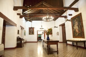 Um corredor amplo com teto alto, vigas de madeira, um candelabro, ventiladores de teto e uma mesa com um vaso de flores.
