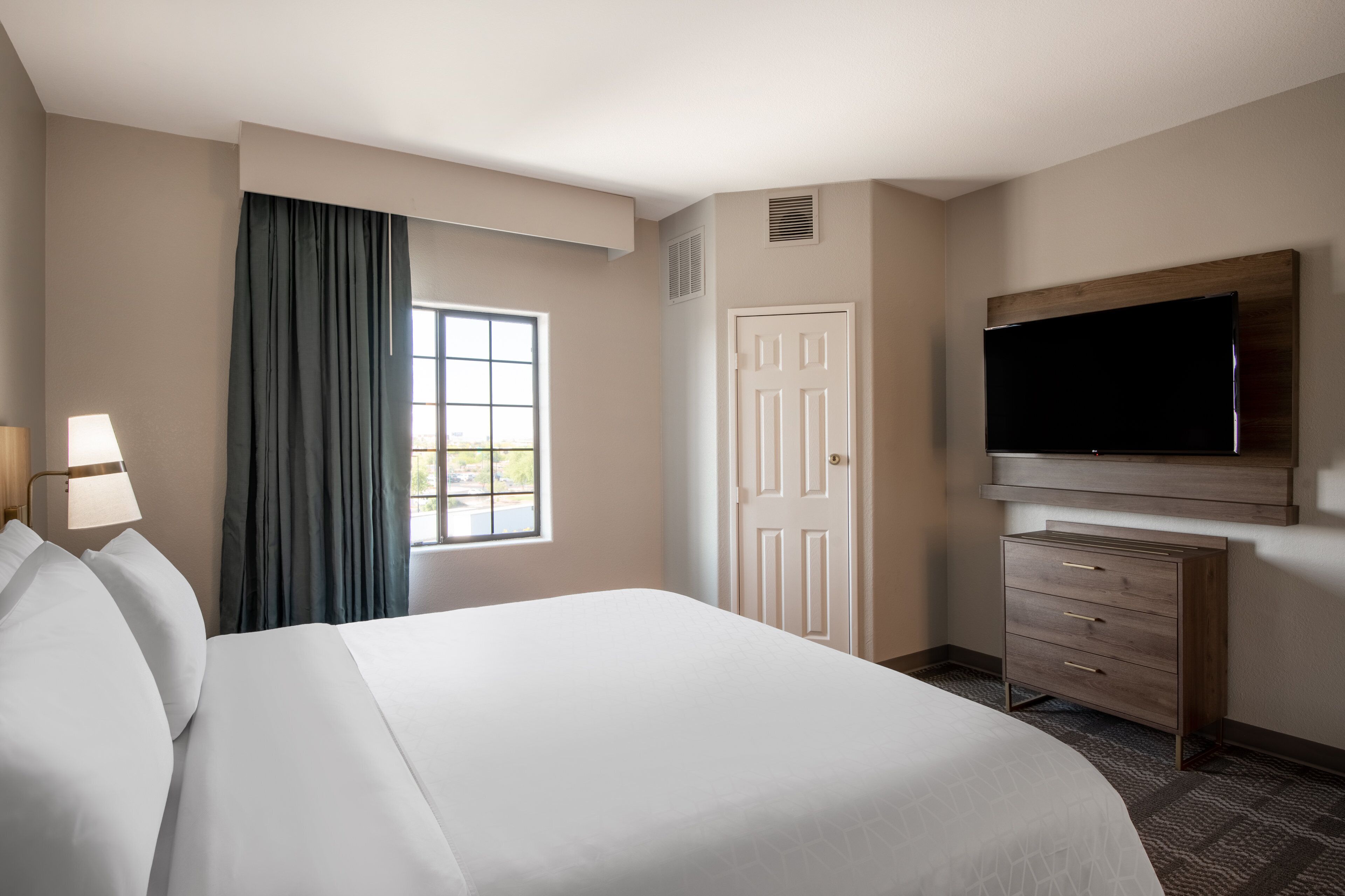 Quarto de hotel com uma cama grande, televisão de tela plana na parede, cômoda, janela com cortinas e luminária na mesinha de cabeceira.