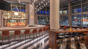Uma área de bar moderna com balcão circular, vários bancos e uma grande janela com vista para a cidade.