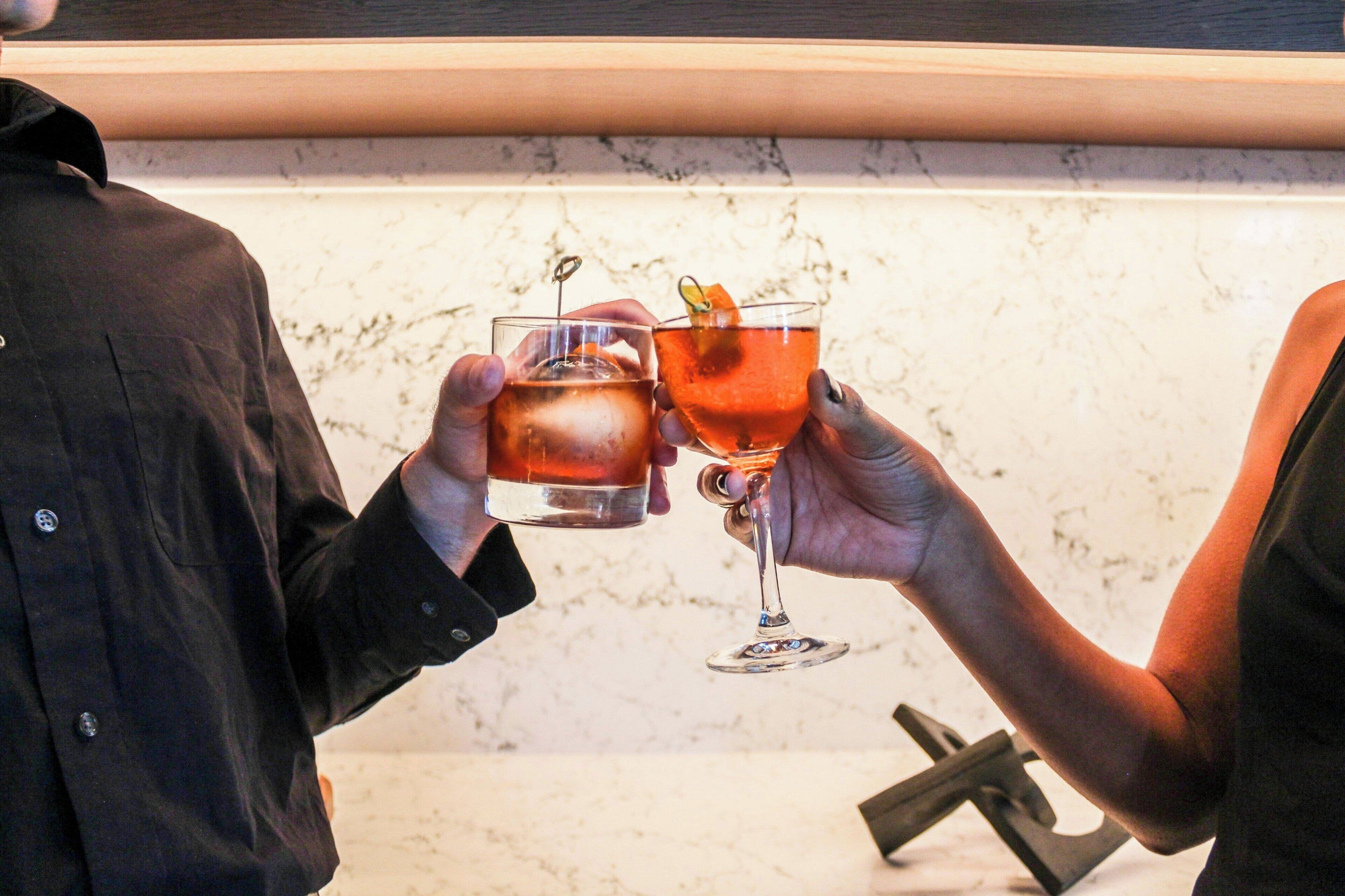 Duas pessoas segurando copos com uma bebida vermelha, provavelmente um cocktail, gelo e guarnições.