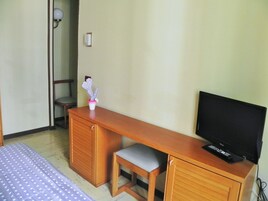 Um quarto pequeno e simples, com uma escrivaninha de madeira, uma televisão e uma cama com colcha de bolinhas.