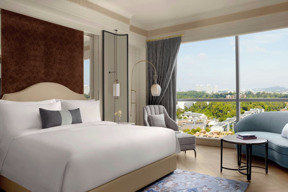 Quarto de hotel moderno com uma cama grande, área de estar com sofá e poltronas, uma mesinha e uma janela ampla com vista para a cidade.