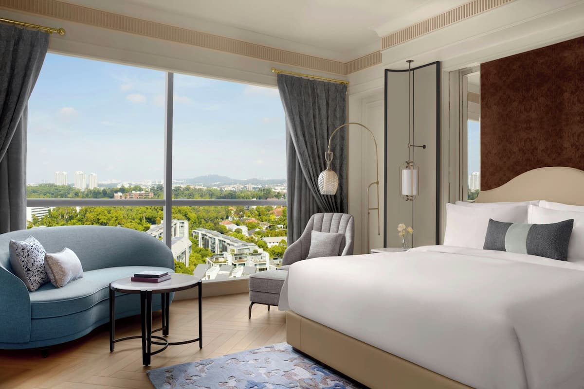 Quarto de hotel com uma cama grande, um sofá curvo, uma mesa pequena e vista para a paisagem urbana através de amplas janelas.