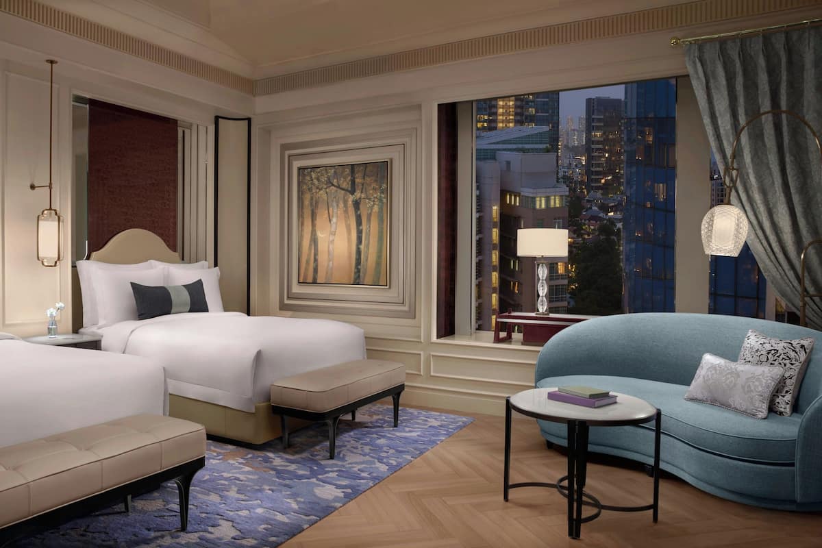 Quarto de hotel com duas camas, um sofá, uma mesinha e vista para a cidade à noite.