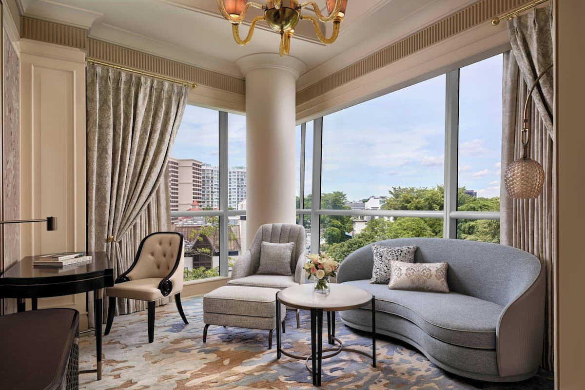 Quarto de hotel moderno com uma janela ampla, um sofá cinzento, uma poltrona combinando, uma mesa pequena e um vaso decorativo.