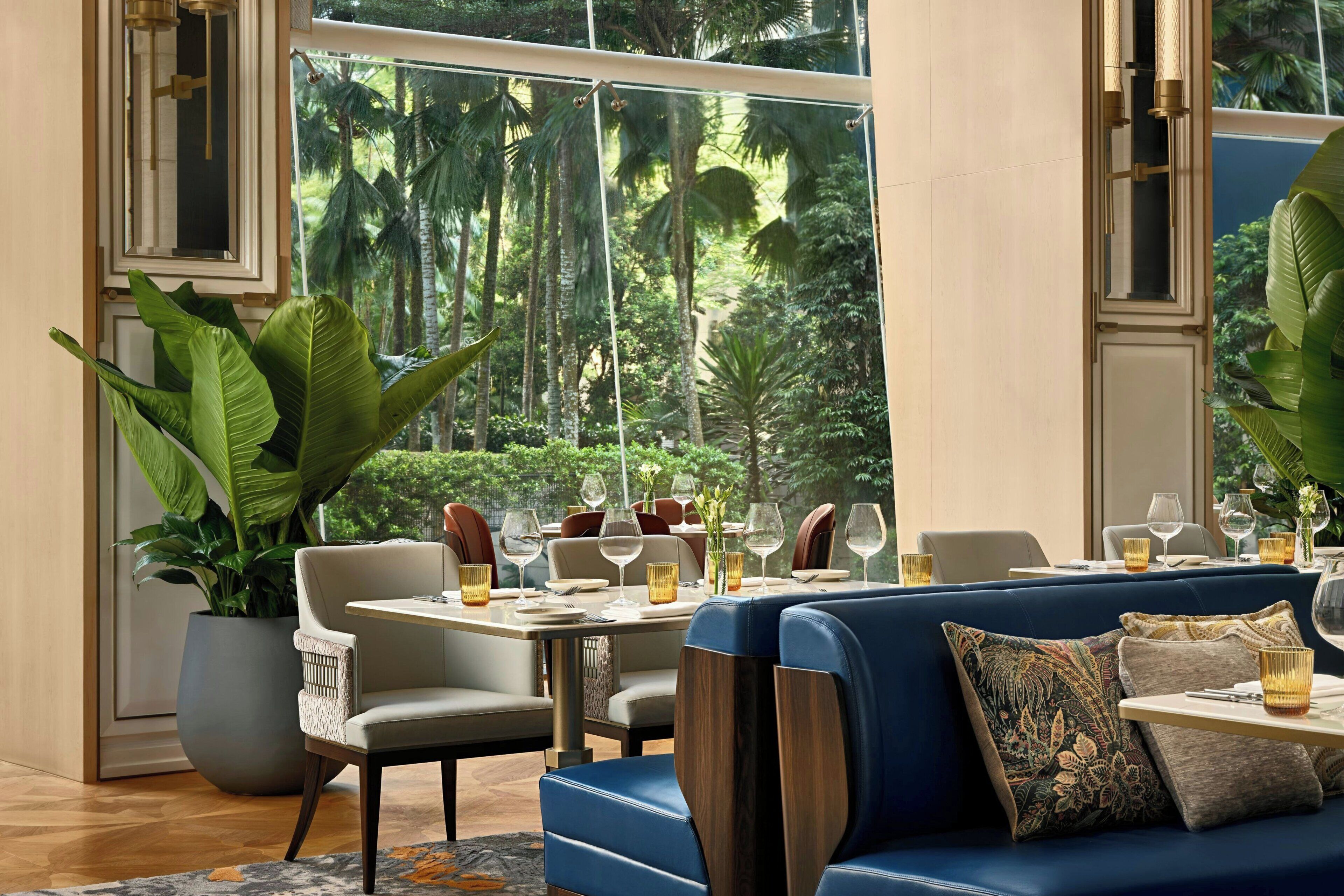 Um restaurante com vista para árvores tropicais, plantas em vasos grandes e um sofá azul com almofadas estampadas.