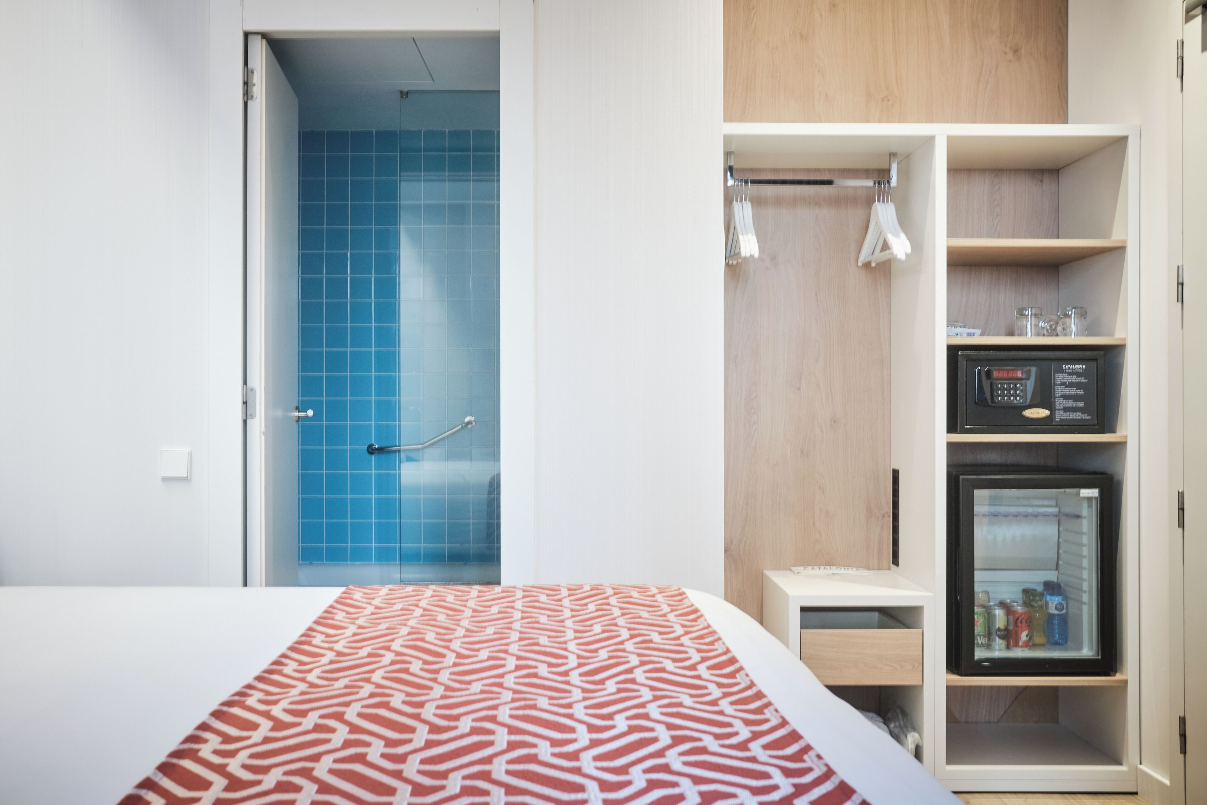 Quarto de hotel com cama, banheiro com azulejos azuis, armário com prateleiras e um minibar.