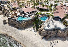 Vista aérea de um resort costeiro com várias piscinas, imóveis na orla e palmeiras.
