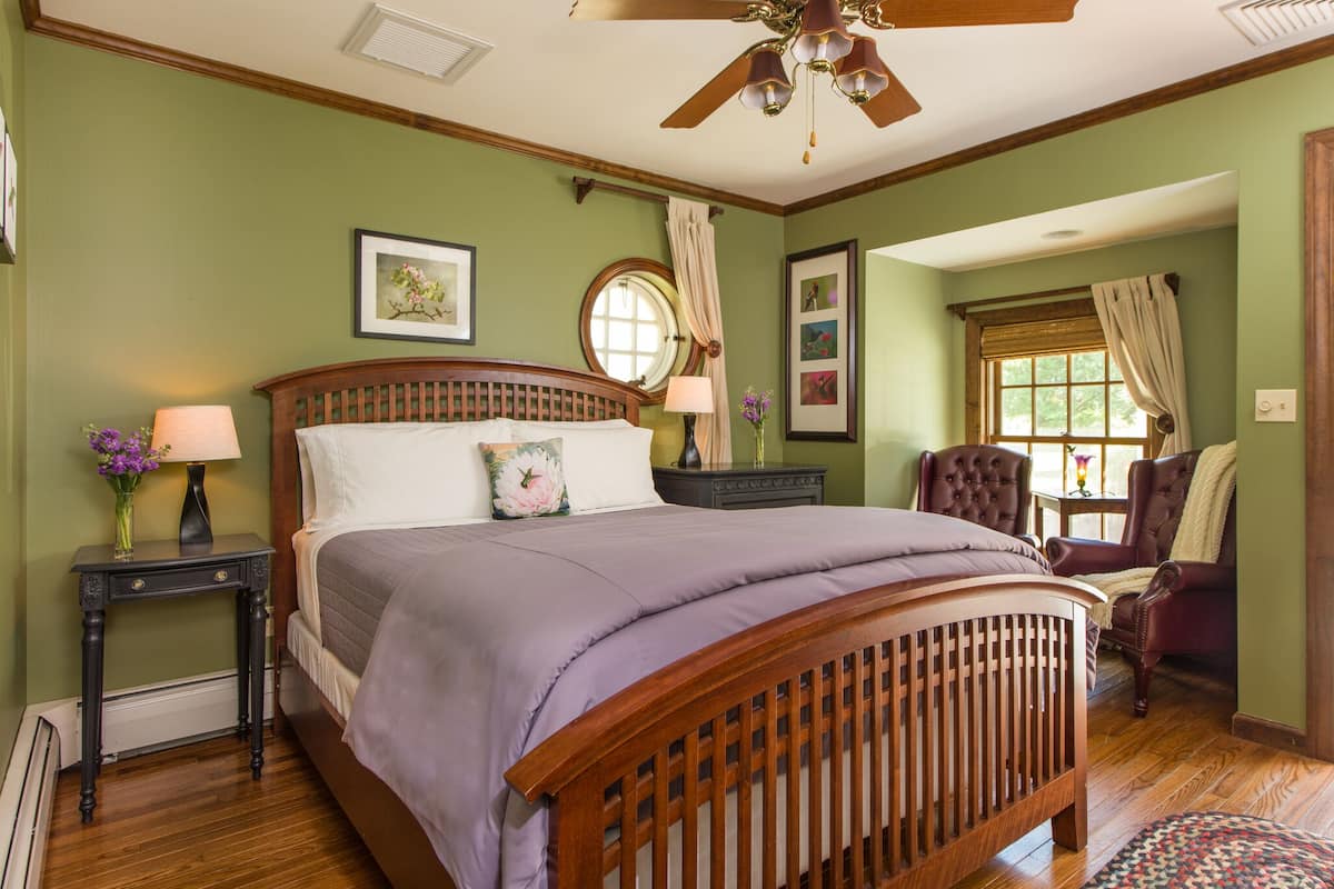 Quarto com cama de madeira, mesa de cabeceira com luminária, cadeira e janela com cortinas.