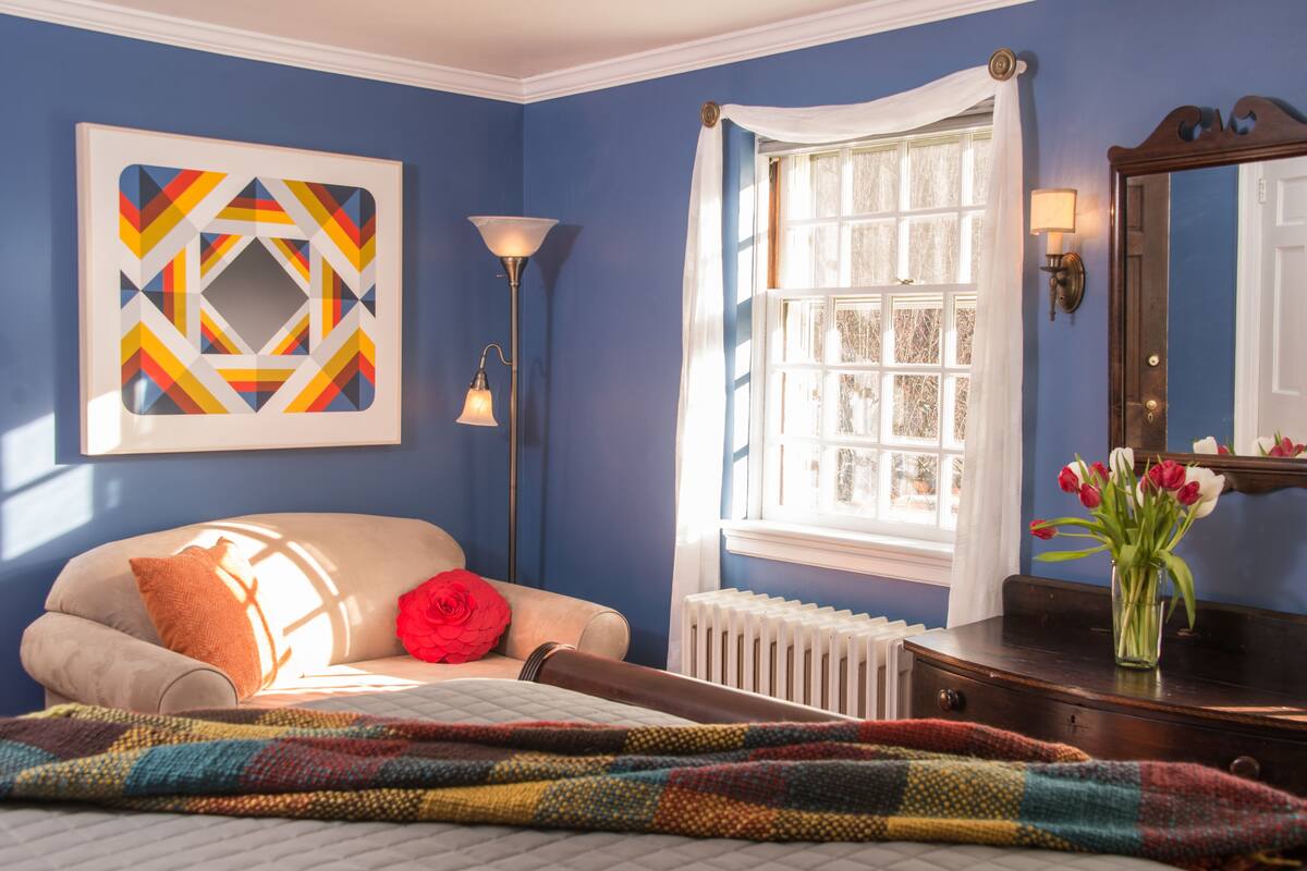 Quarto com cama, janela com cortinas brancas, radiador, abajur, espelho e um vaso com flores.