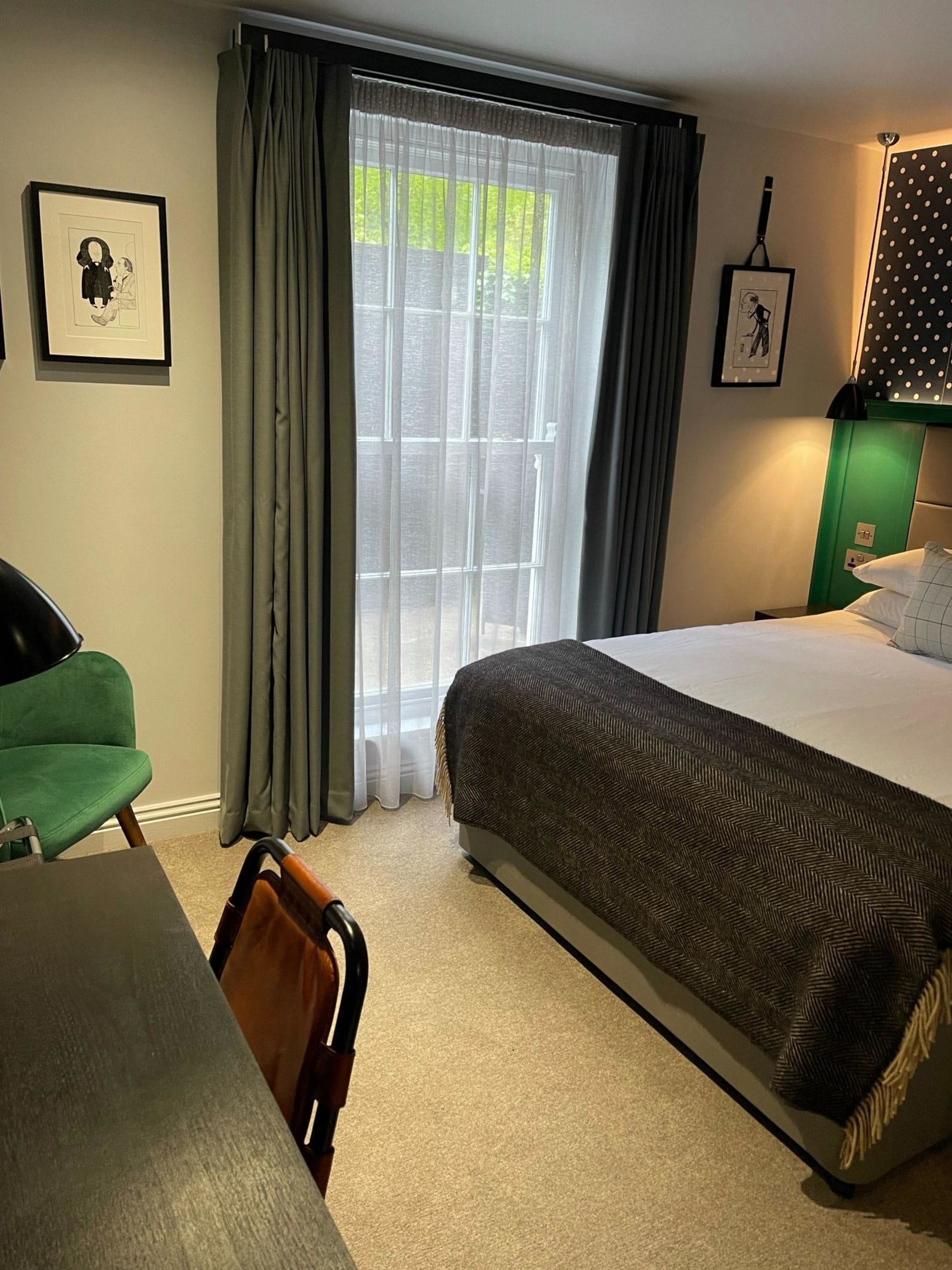 Quarto de hotel com cama, escrivaninha, cadeira, janela com cortinas e obras de arte nas paredes.
