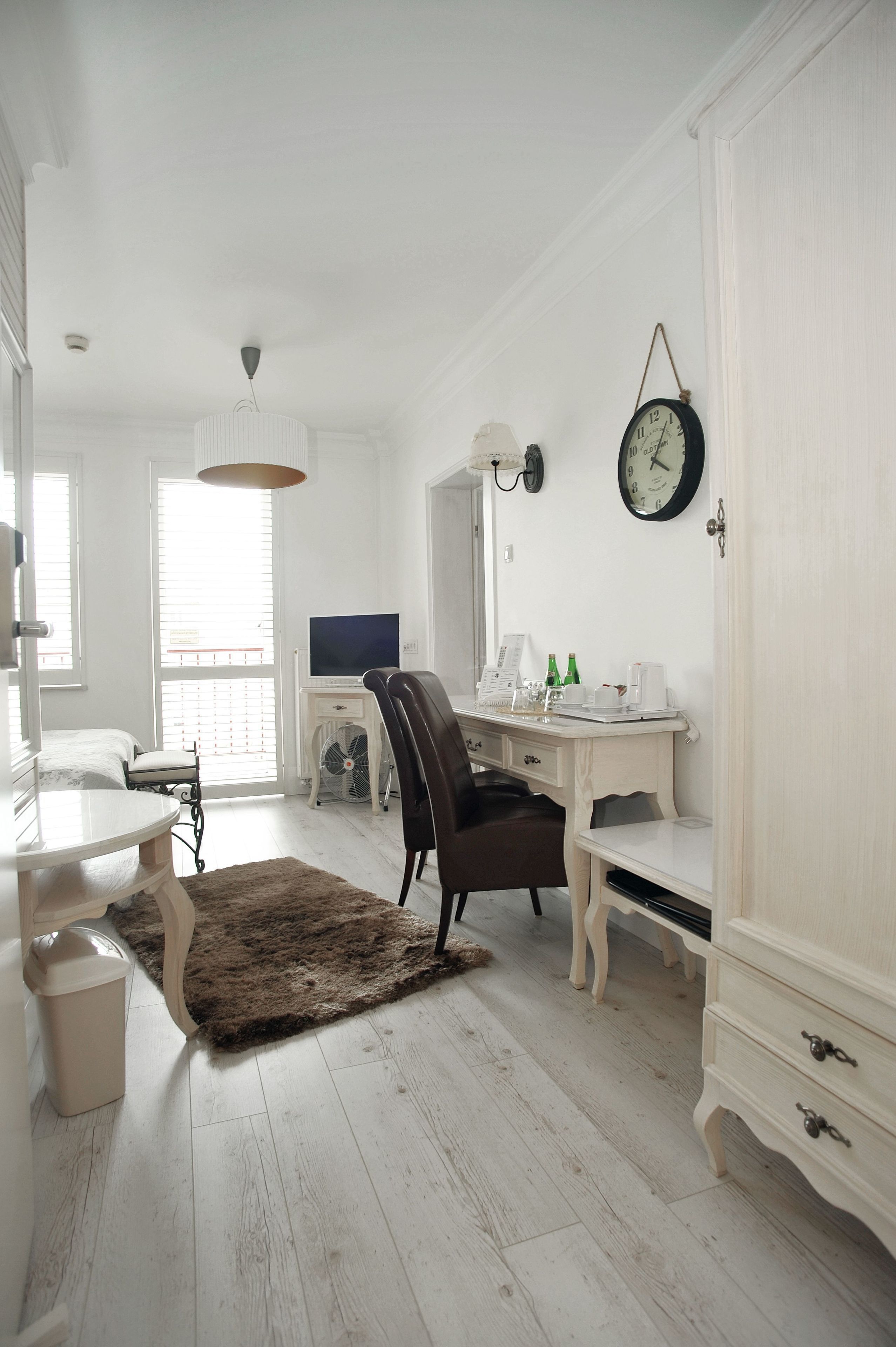 Um quarto espaçoso com decoração em tons de branco, com uma escrivaninha, uma cadeira, uma televisão e um relógio na parede.