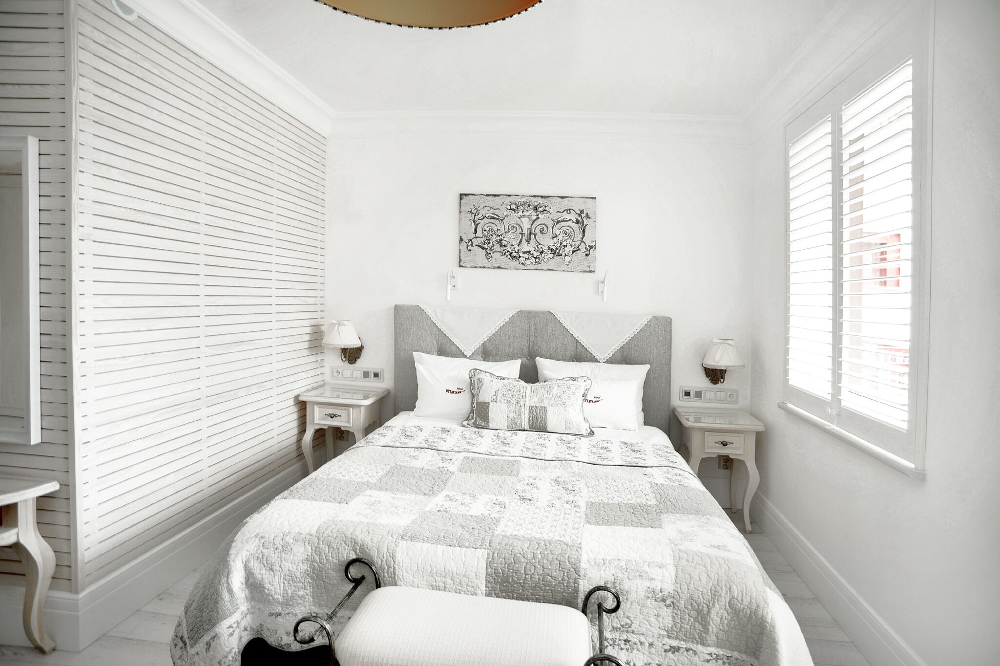 Quarto com cama, duas mesas de cabeceira, um criado-mudo, janela com persianas e uma obra de arte na parede.