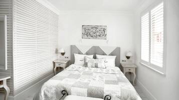 Quarto com cama, duas mesas de cabeceira, um criado-mudo, janela com persianas e uma obra de arte na parede.