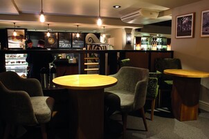Um café acolhedor com mesas de madeira redondas e cadeiras estofadas, uma área de bar com garrafas e um quadro de cardápio.