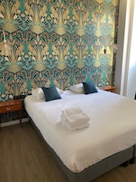 Quarto de hotel com uma cama grande, duas mesas de cabeceira e uma parede com papel de parede estampado com motivos florais.