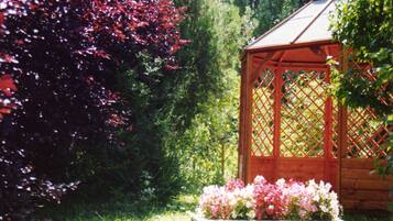 Um gazebo de madeira com telhado treliçado, cercado por vegetação exuberante e plantas floridas.