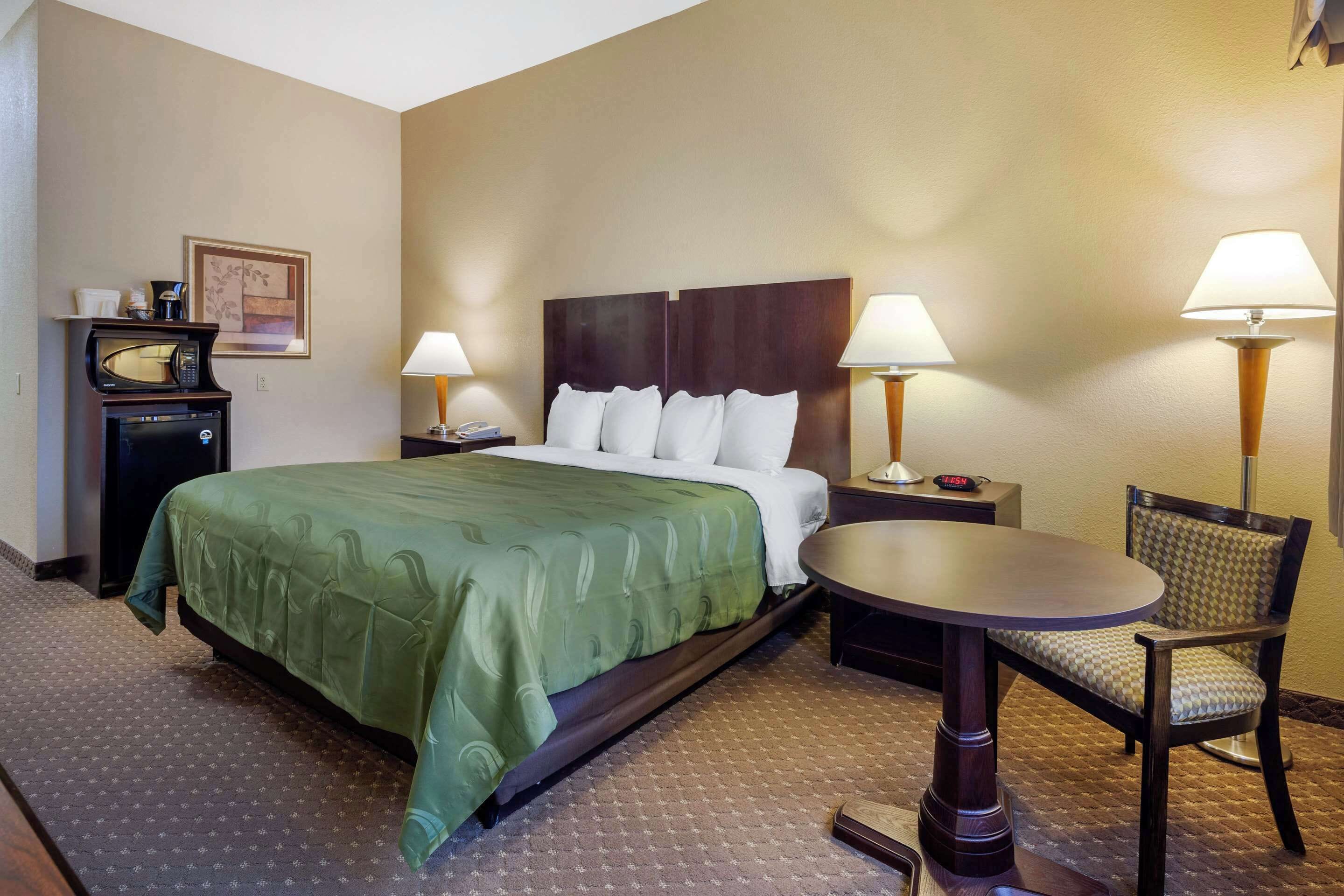 Quarto de hotel com uma cama, duas luminárias de cabeceira, uma mesa, uma cadeira, micro-ondas e uma pequena prateleira com objetos.
