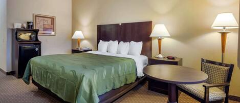 Quarto de hotel com uma cama, duas luminárias de cabeceira, uma mesa, uma cadeira, micro-ondas e uma pequena prateleira com objetos.