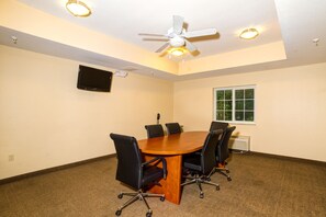 Uma sala de conferências com uma mesa de madeira, quatro cadeiras de escritório pretas, um ventilador de teto, uma TV de tela plana fixada na parede e uma janela com persianas.