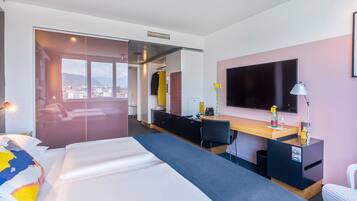 Quarto de hotel com uma cama grande, uma escrivaninha, uma cadeira, uma TV e vista para a cidade.