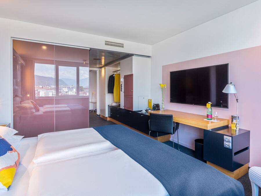 Quarto de hotel com uma cama grande, uma escrivaninha, uma cadeira, uma TV e vista para a cidade.