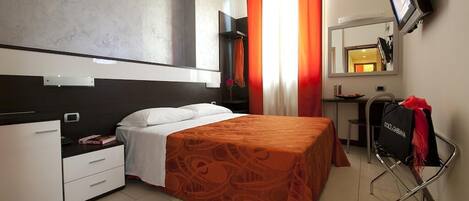 Quarto de hotel com cama, escrivaninha, cadeira, televisão e janela com cortinas.