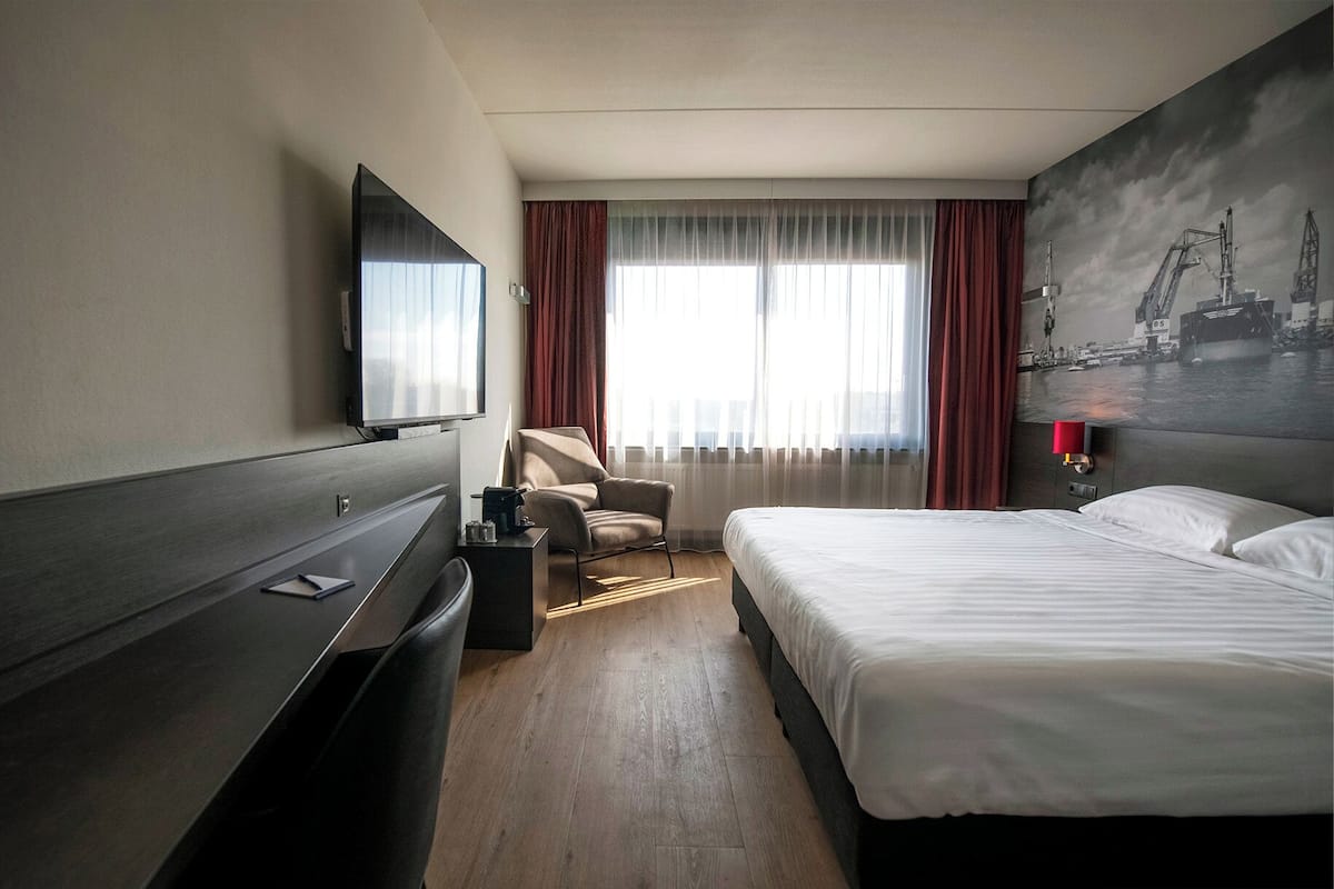 Quarto de hotel com uma cama, escrivaninha, cadeira, televisão e uma grande janela com cortinas.