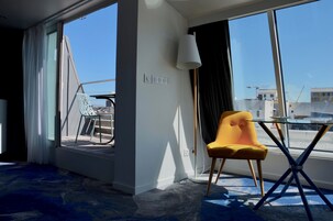 Quarto de hotel com uma grande janela, uma cadeira amarela, uma mesa de vidro e um abajur.