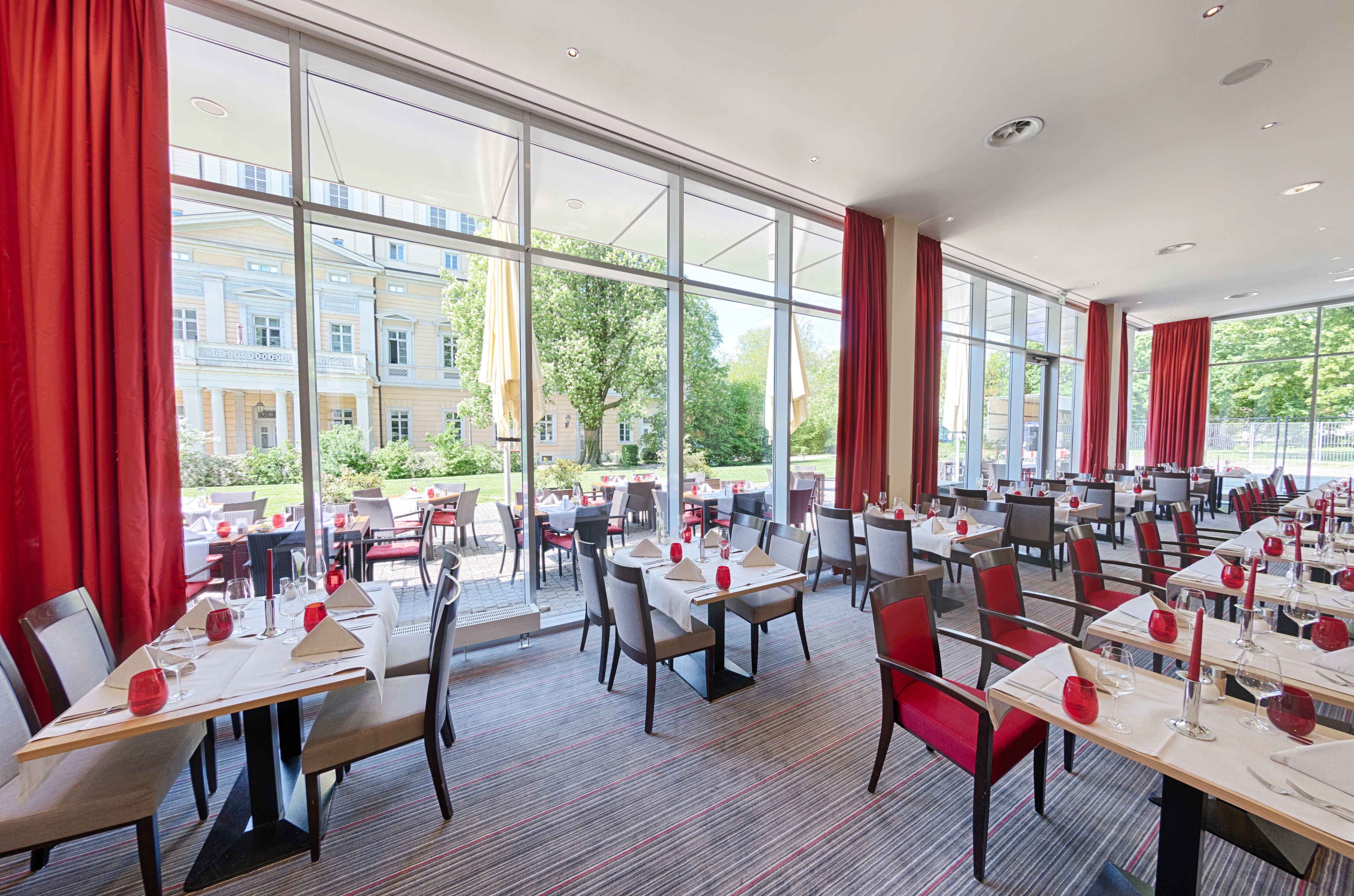 Restaurante com várias mesas e cadeiras, grandes janelas e cortinas vermelhas.