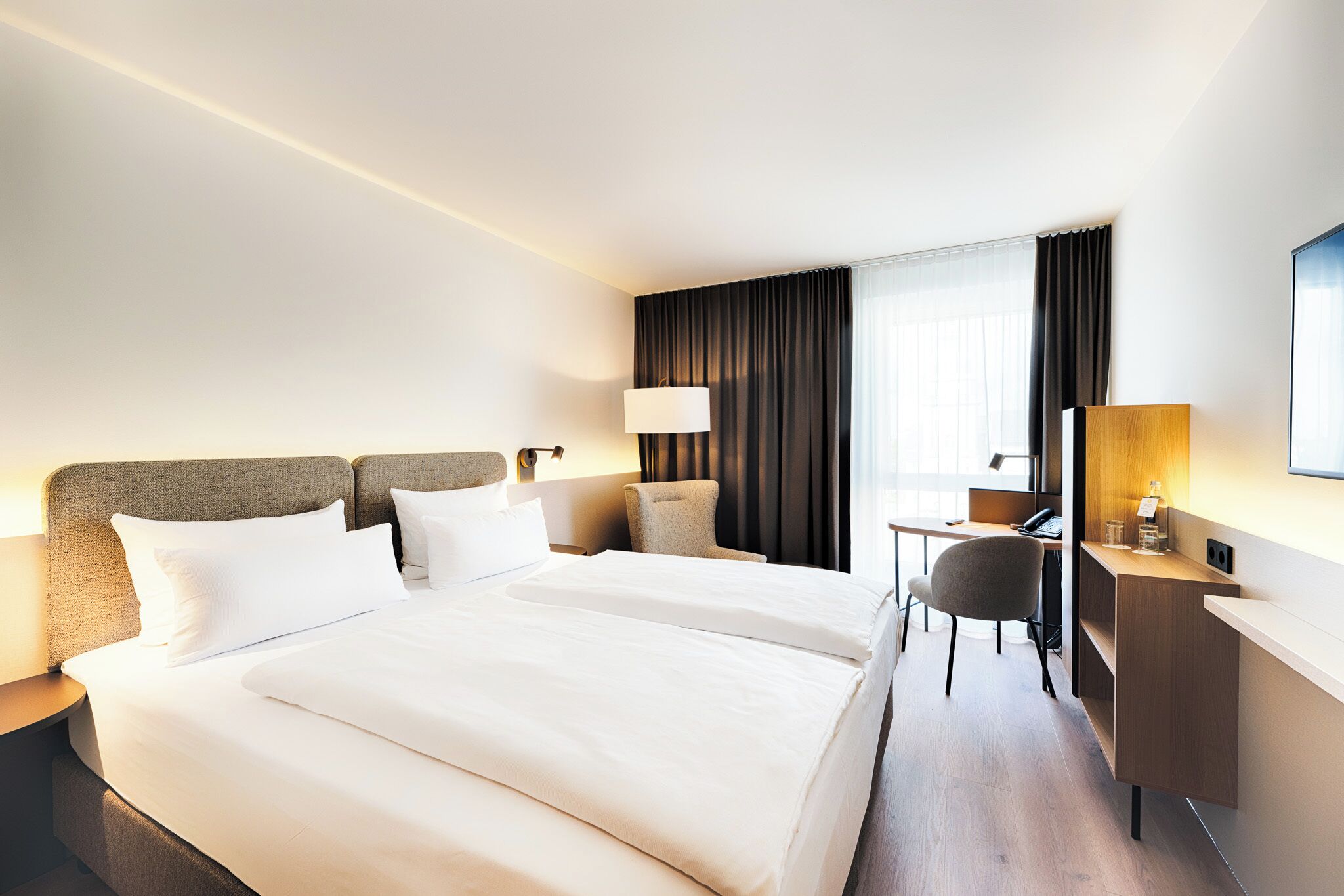 Quarto de hotel com uma cama grande, uma escrivaninha, uma cadeira e uma luminária.