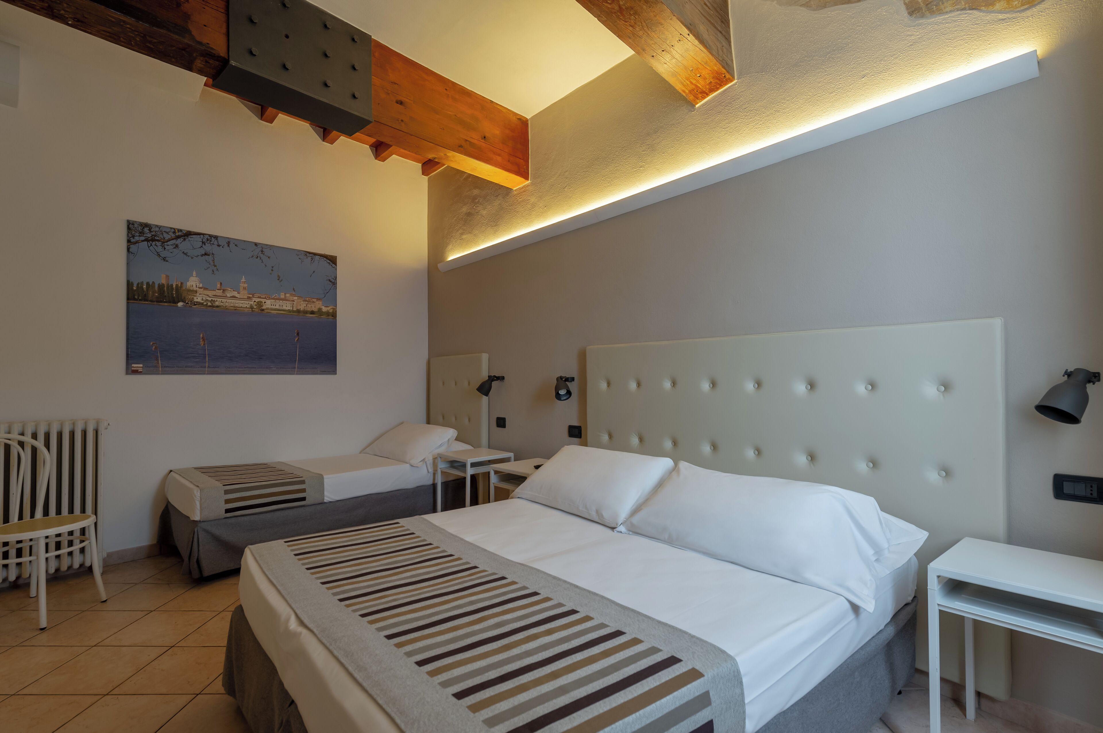 Quarto de hotel com uma cama grande, duas camas menores, uma cadeira, uma escrivaninha e um quadro de uma paisagem urbana.