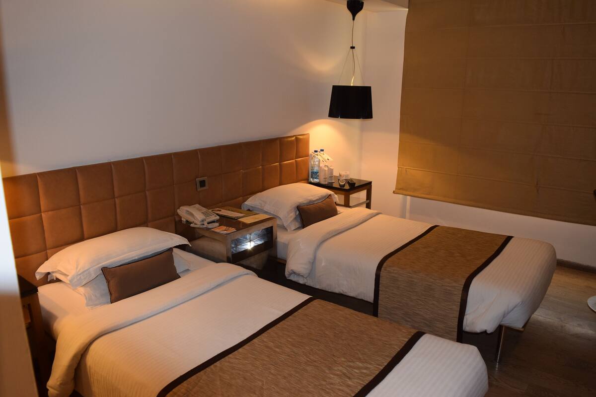 Quarto de hotel com duas camas, uma mesa de cabeceira com telefone e produtos de higiene, luminária fixada na parede e cabeceira de madeira.