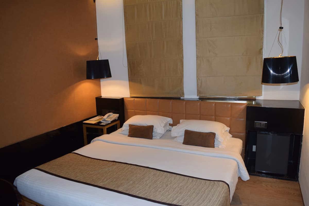 Quarto de hotel com uma cama grande, uma mesinha de cabeceira com telefone, um pequeno frigorífico preto e duas luminárias fixadas na parede.