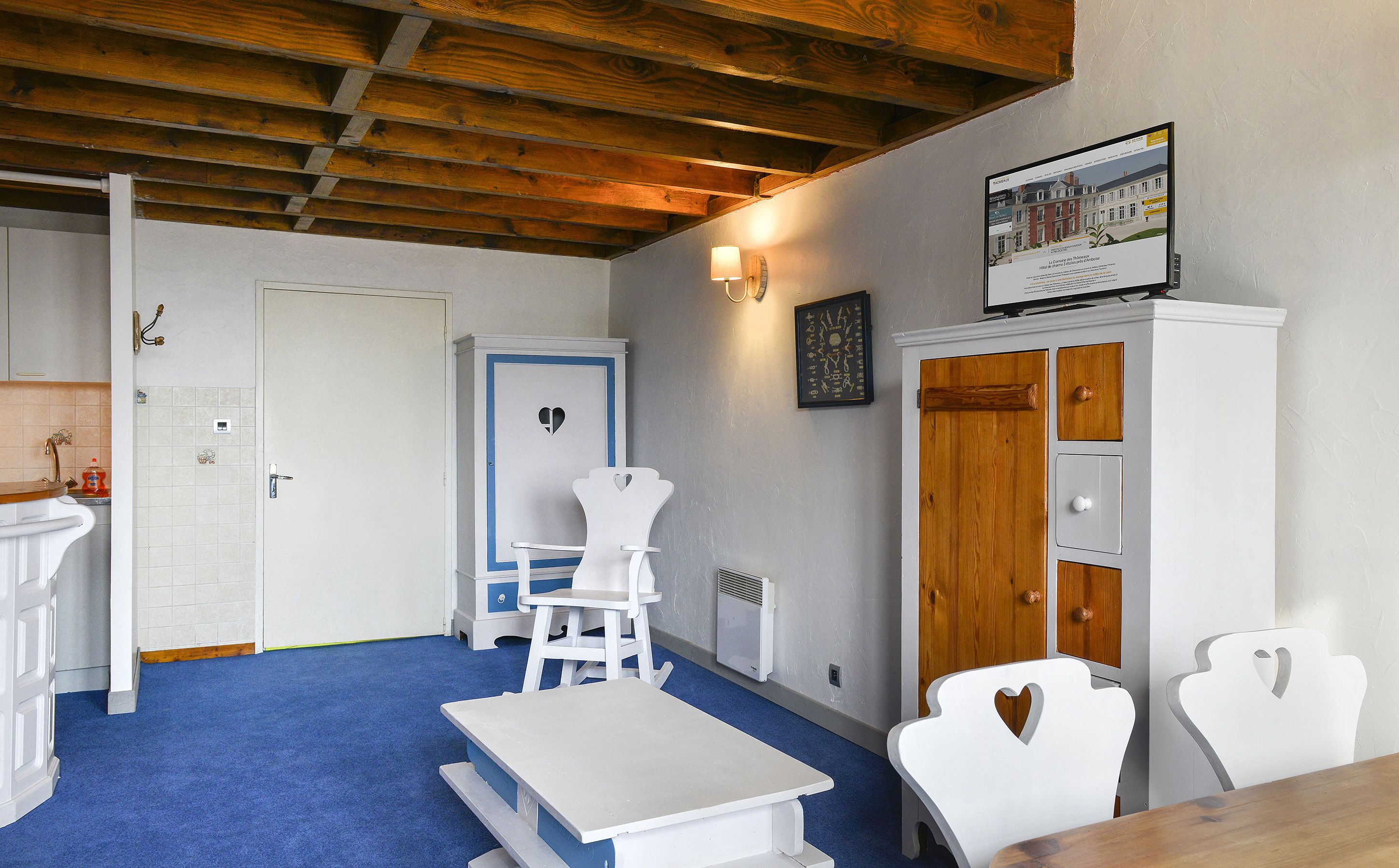Um quarto com teto de madeira, porta branca com recorte em formato de coração, mesa branca e cadeira branca com recorte em formato de coração.