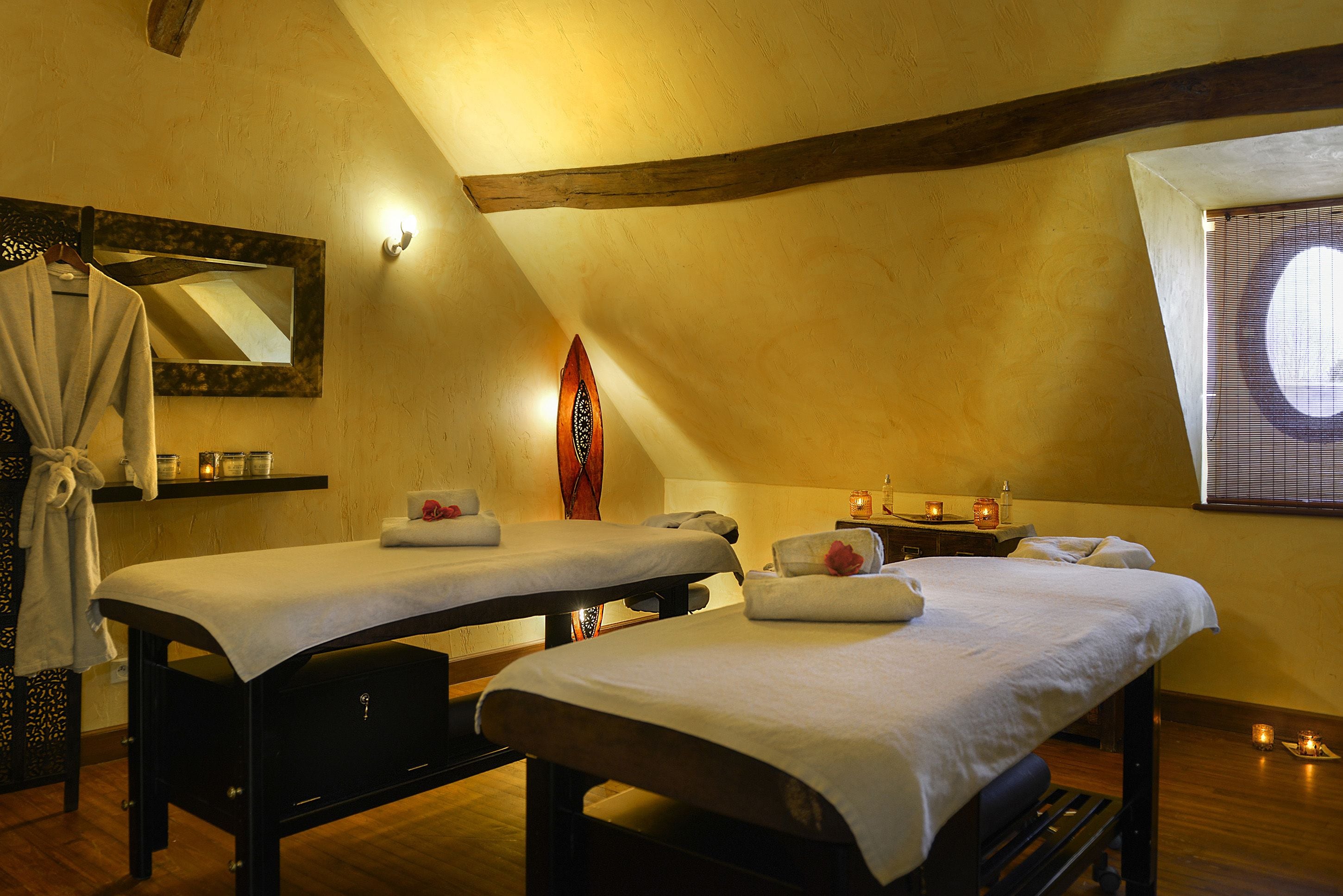 Um quarto com duas mesas de massagem, um espelho, uma janela com persianas e uma pequena estante com velas.