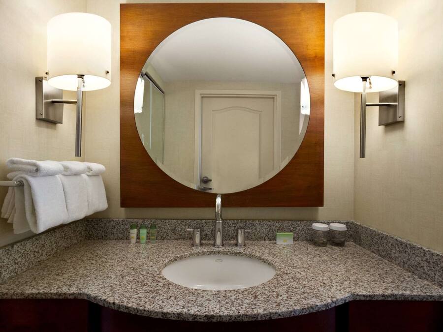 Uma casa de banho com bancada de granito, um espelho redondo, duas luzes na parede, um porta-toalhas e uma porta.